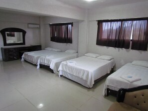 Quarto de hotel com três camas de solteiro, uma escrivaninha pequena, um espelho e uma televisão.