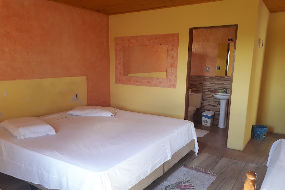 Quarto com cama, banheiro pequeno com pia e espelho, e porta que dá para outro cômodo.