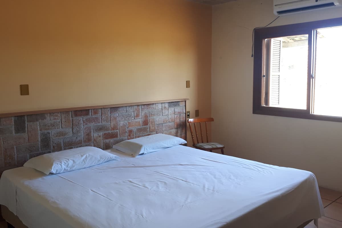 Uma cama com roupa de cama branca, cabeceira em padrão de tijolos e uma cadeira de madeira ao lado.