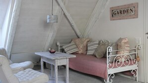 Um quarto de sótão acolhedor com uma cama de metal branca, uma mesinha e uma cadeira. Uma placa com a inscrição 'GARDEN' é visível na parede.