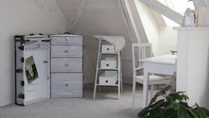 Quarto com teto inclinado, paredes de madeira branca, uma escrivaninha branca com cadeira e uma escada com gavetas.