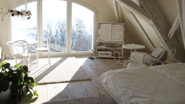 Quarto com uma grande janela, uma cama, uma mesinha, uma cadeira e uma estante de livros.