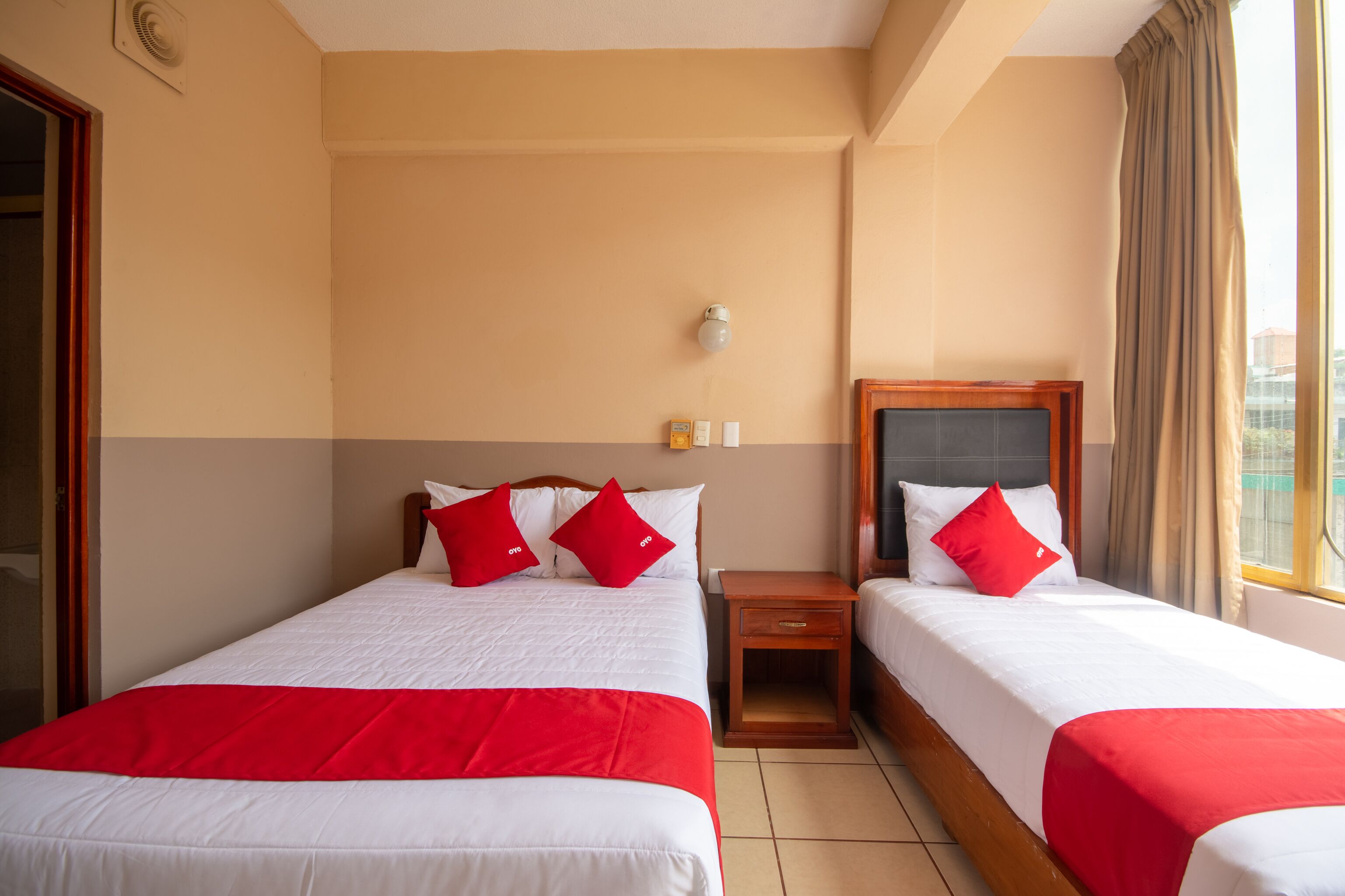 Quarto de hotel com duas camas, cada uma com roupa de cama vermelha e branca, uma mesinha de cabeceira de madeira, uma luminária fixada na parede e uma janela com cortinas.