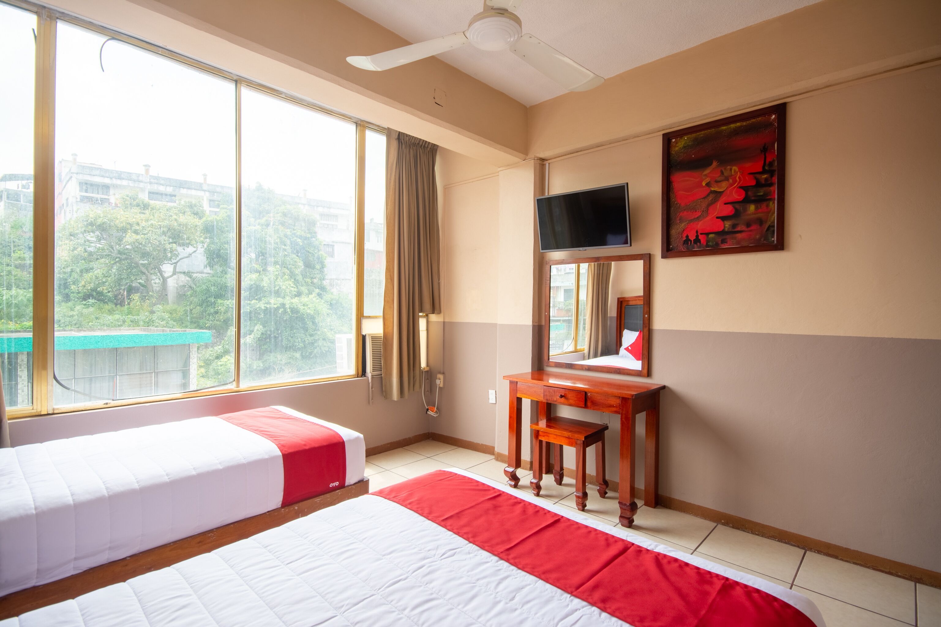 Quarto de hotel com cama, escrivaninha, cadeira, televisão e janela com vista para árvores e edifícios.