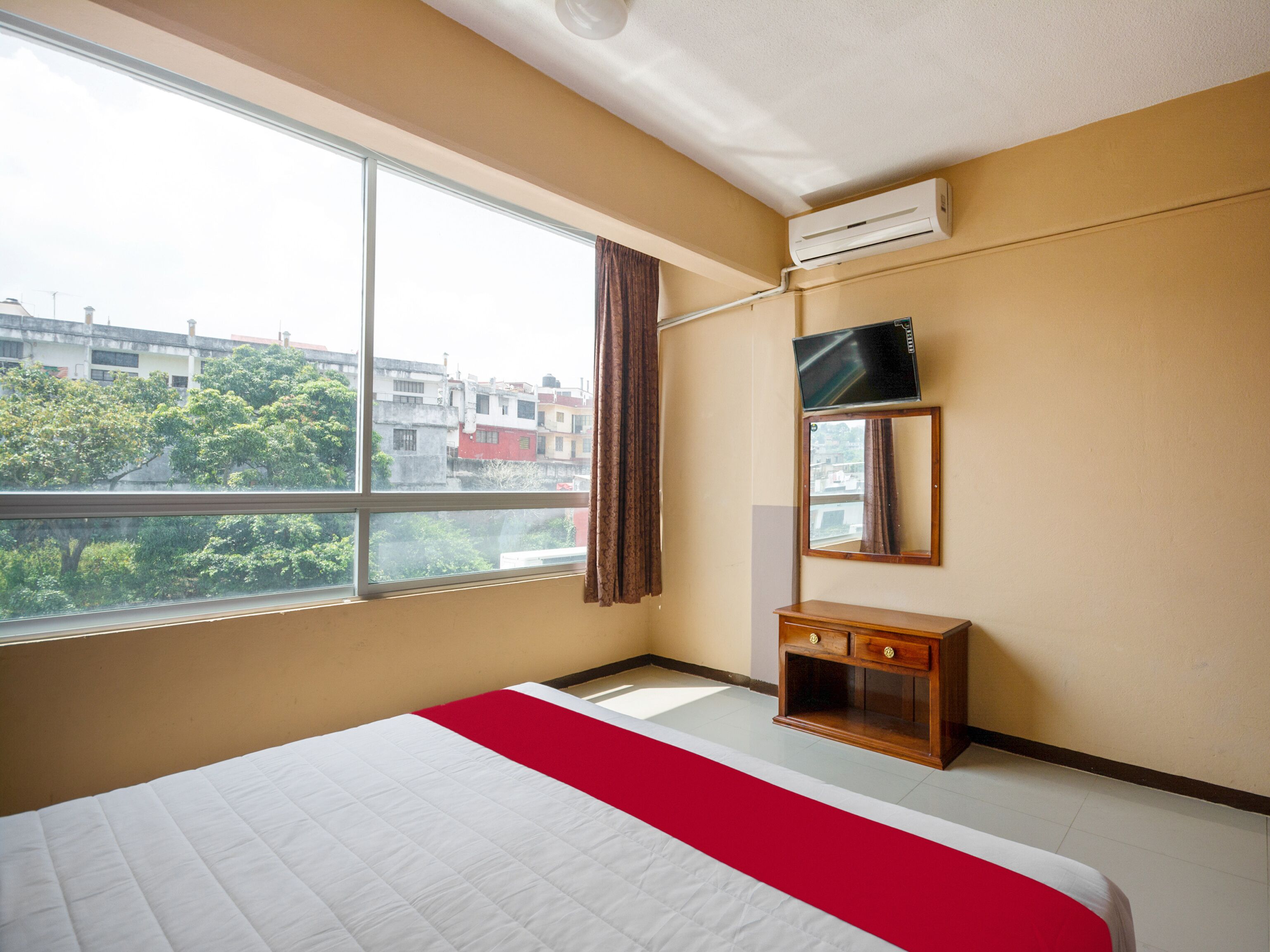Quarto de hotel com uma janela grande, cama com detalhe em vermelho, mesa de cabeceira de madeira e ar condicionado.