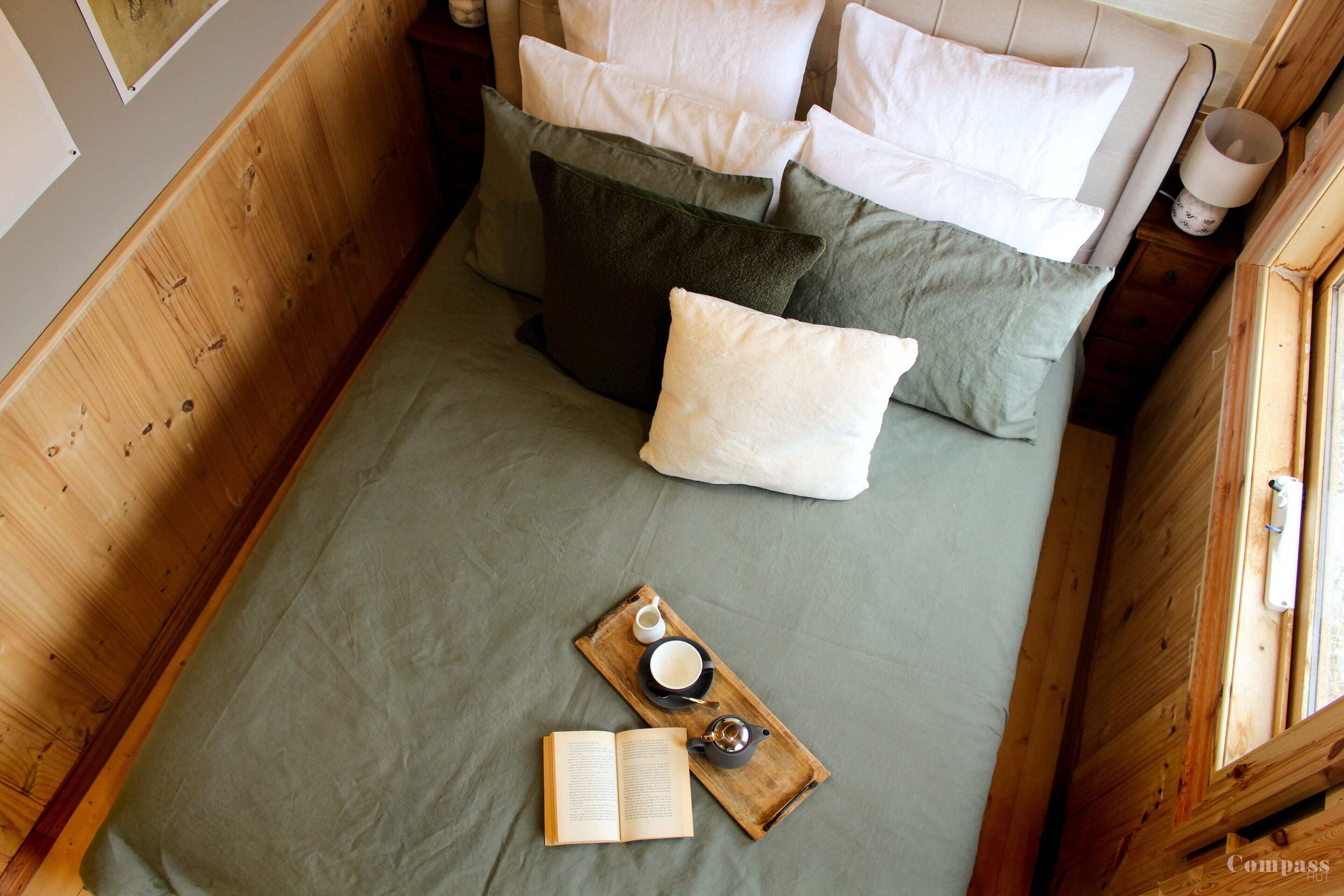 Quarto tipo cabana de madeira com cama, travesseiros, bandeja com chaleira, xícaras e um livro.