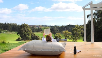 Uma pessoa deitada em um grande travesseiro branco em um deck de madeira, lendo um livro com uma vista panorâmica de uma paisagem verde e árvores ao fundo.