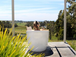 Uma mulher relaxando em uma banheira branca em um deck de madeira, com uma paisagem verde e um corpo d'água ao fundo.