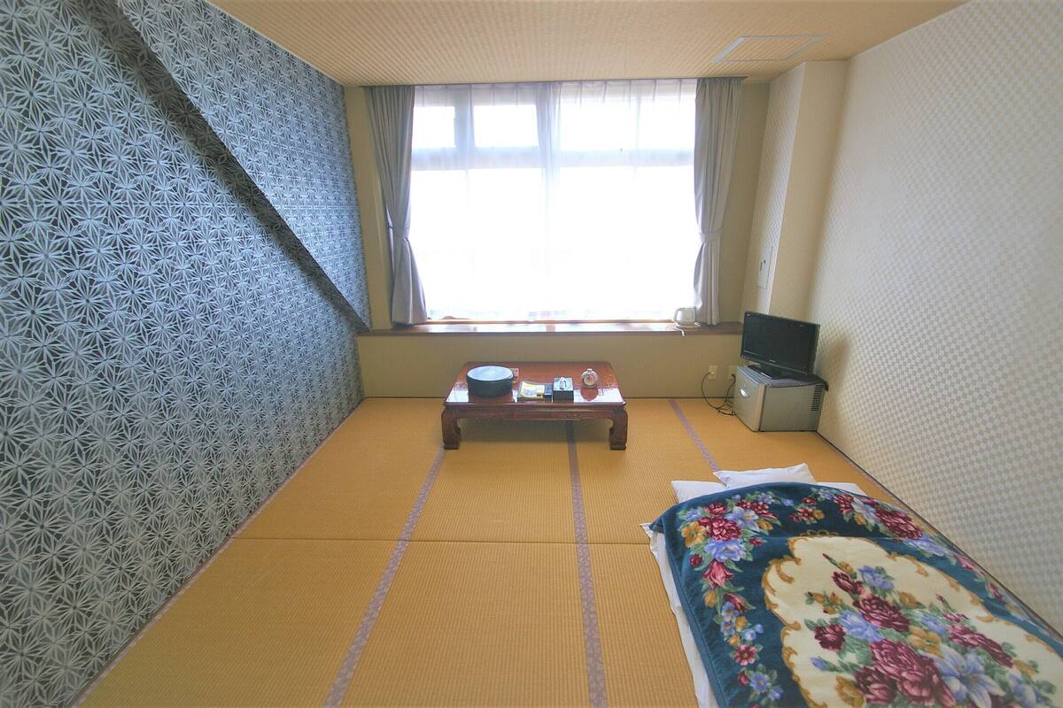 Quarto com piso de tatame, cama futon, uma mesinha com uma tigela e um livro, televisão e uma janela com cortinas.