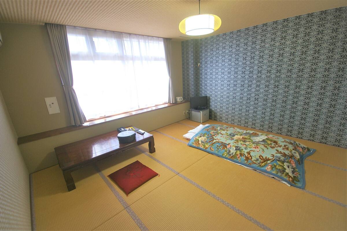 Um quarto japonês tradicional com tatames, futon, uma pequena mesa de madeira e uma televisão.