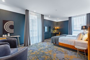 Quarto de hotel com cama, escrivaninha, cadeira, televisão e uma janela grande com cortinas.