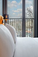 Quarto de hotel com uma janela grande, cama com roupa de cama branca, abajur preto e vista para a paisagem urbana.