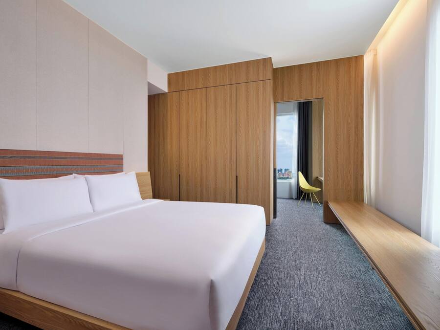 Quarto de hotel moderno com uma cama bem arrumada, guarda-roupa de madeira e uma pequena área de estar com uma cadeira.