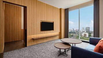 Quarto de hotel moderno com uma janela grande que oferece vista para a cidade, uma TV de tela plana fixada na parede e uma área de estar com duas mesas redondas.