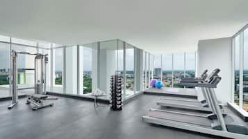 Academia moderna com esteiras, aparelhos de musculação e saco de pancada, com grandes janelas e vista para a cidade.