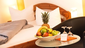 Uma cama com cabeceira de madeira, uma tigela de frutas sobre uma mesinha ao lado, e uma placa com um nome.