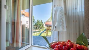 Quarto com porta de vidro deslizante que dá para um parquinho, uma tigela de cerejas e vista de um edifício com telhado vermelho.