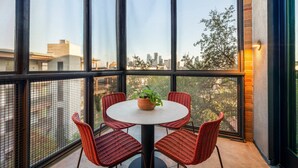 Mesa redonda com quatro cadeiras vermelhas e um vaso de planta no centro, com vista para o horizonte urbano através de grandes janelas.