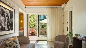 Uma sala de estar aconchegante com teto de madeira, uma grande moldura com imagem, um sofá com almofadas estampadas, duas poltronas, uma mesa de centro e vista através de uma porta de vidro para uma área de estar externa.