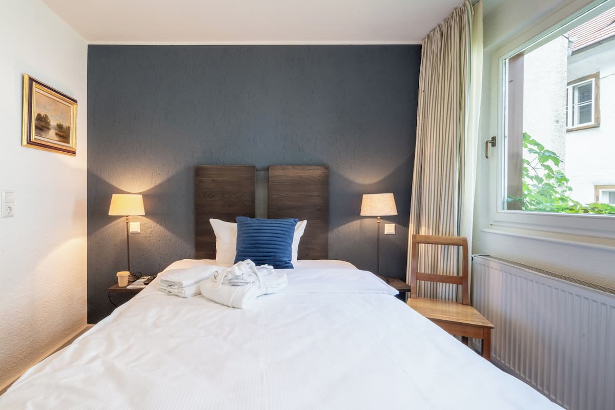 Cama bem arrumada com lençóis brancos e travesseiro azul, duas luminárias de cabeceira, uma cadeira de madeira e uma janela com cortinas.