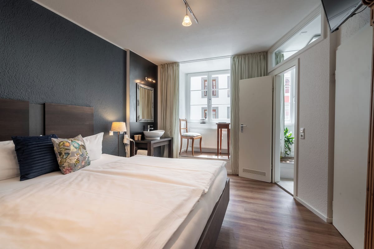 Quarto de hotel com uma cama grande, uma mesa de cabeceira com abajur, uma janela com cortinas e vista para o exterior.