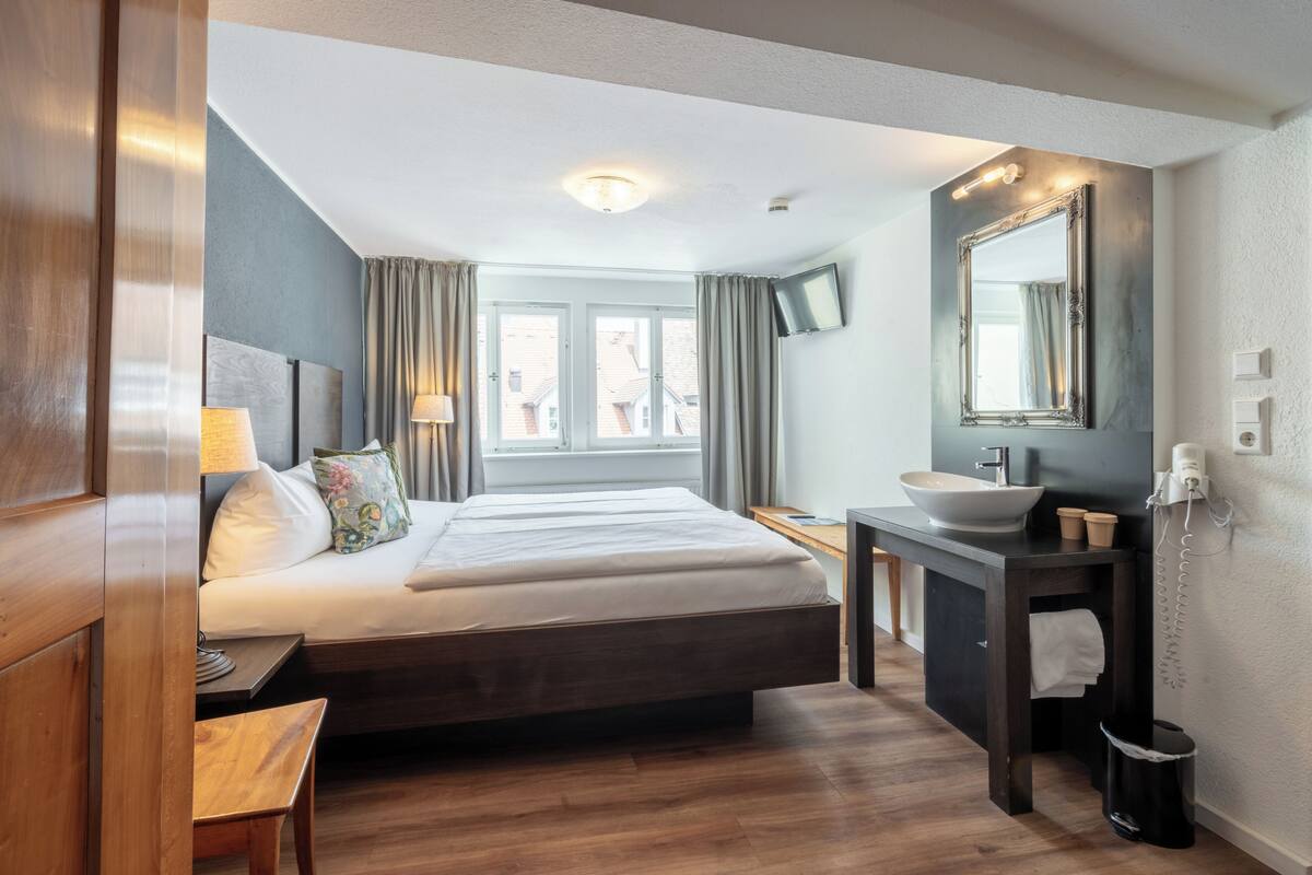 Quarto de hotel moderno com uma cama grande, piso de madeira, mesa de cabeceira com abajur, banheiro com pia e espelho, e vista para o exterior pela janela.