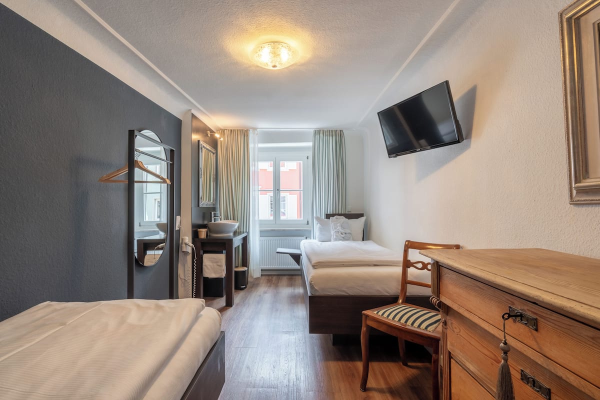 Quarto de hotel com duas camas, cômoda de madeira, televisão e janela com cortinas.