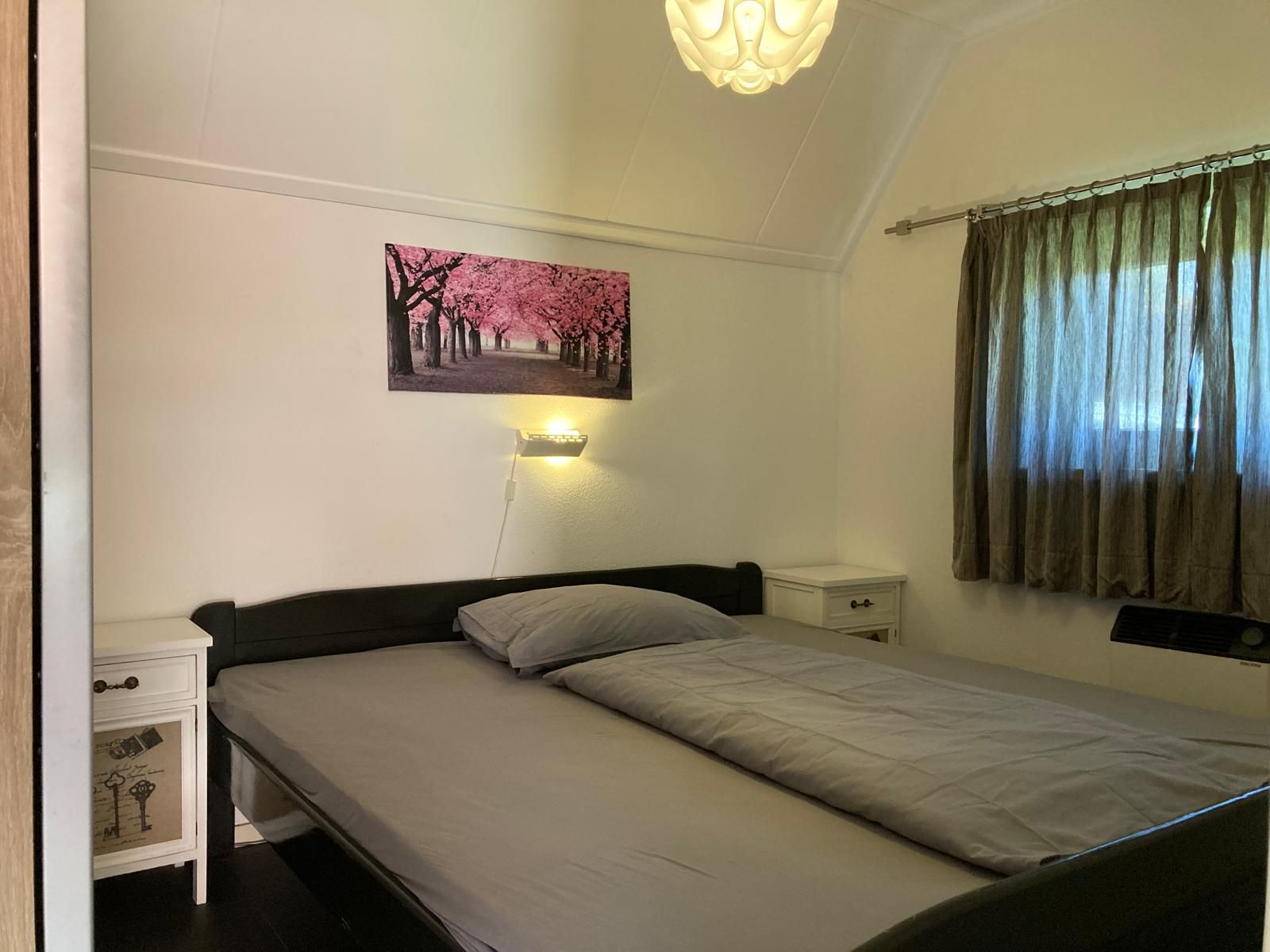 Uma cama de solteiro com cabeceira, mesa de cabeceira, luminária fixada na parede, janela com cortinas e um quadro na parede.