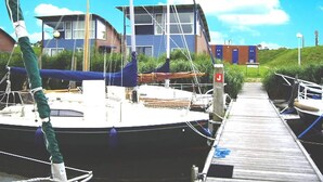 Uma marina com barcos atracados, um cais de madeira e um edifício moderno com fachada azul ao fundo.