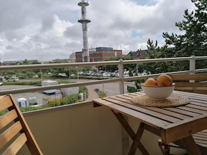 Uma varanda com mesa e cadeiras de madeira, uma cesta de frutas e vista para uma torre e edifícios ao longe.