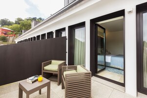 Quarto de hotel moderno com varanda, uma cama com colcha azul e duas cadeiras de madeira com almofadas.