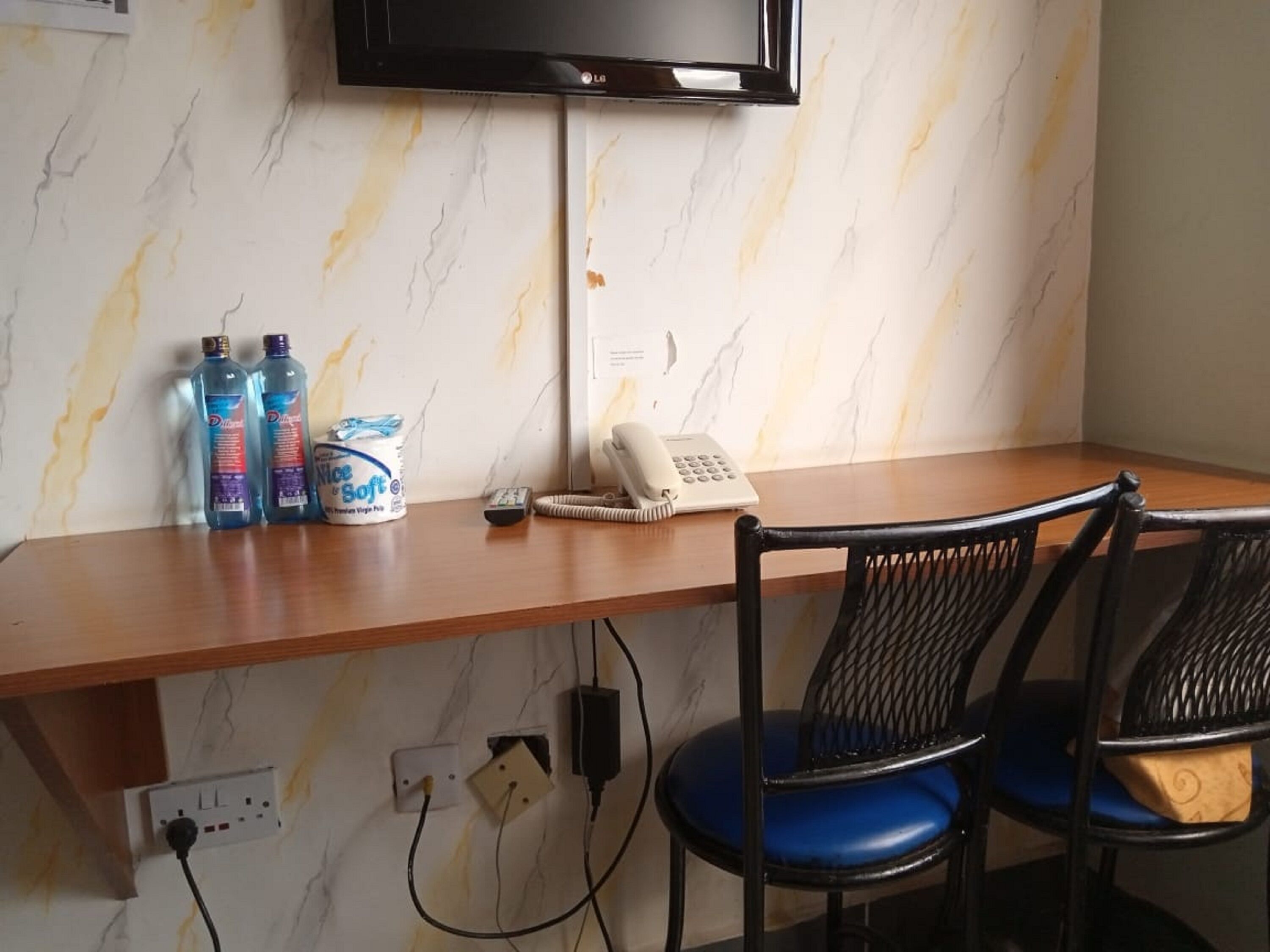Quarto de hotel com escrivaninha de madeira, duas cadeiras pretas, TV fixada na parede, telefone e artigos de higiene no balcão.