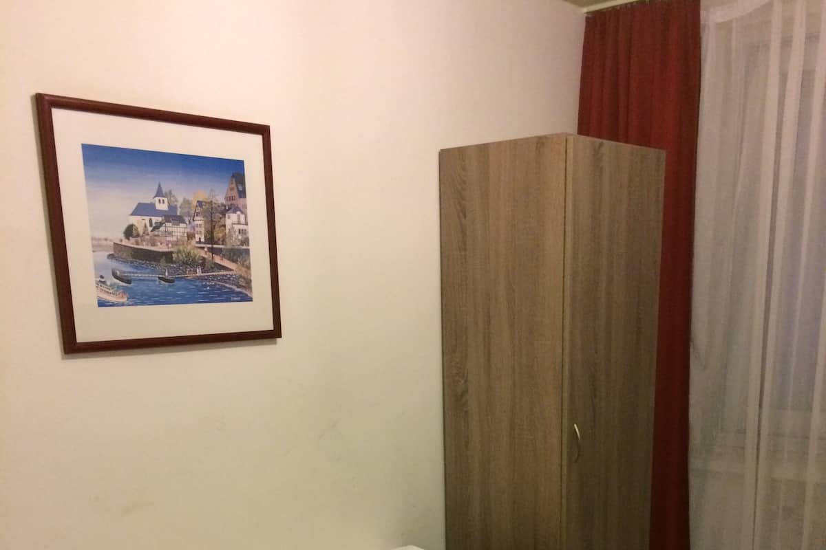 Uma cama de solteiro com colchão branco e manta bege, um guarda-roupa de madeira e um quadro na parede com uma imagem de uma cidade.