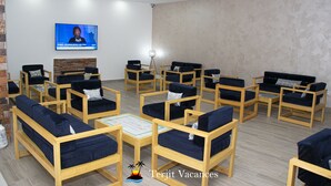 Um quarto com várias cadeiras e sofás de madeira dispostos em formato de U, uma televisão fixada na parede e uma parede de tijolos em um dos lados.
