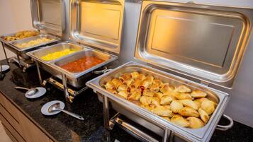 Buffet com diversos alimentos em recipientes metálicos, incluindo aparentemente ovos mexidos, molho vermelho e doces.