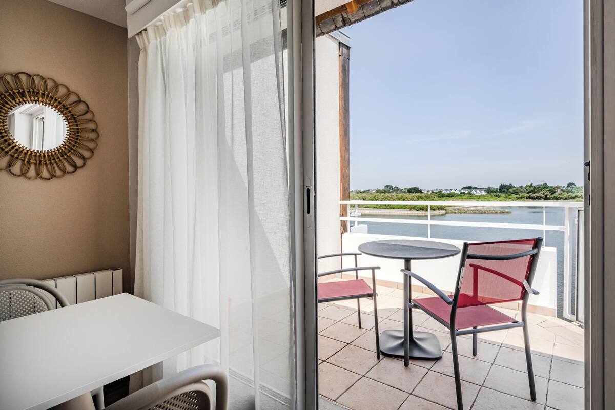 Uma varanda com mesa e cadeiras, vista para um corpo d'água e um espelho decorativo na parede.
