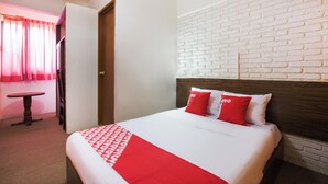 Quarto de hotel com cama, cabeceira de madeira, parede com padrão de tijolos, uma mesinha e uma porta.