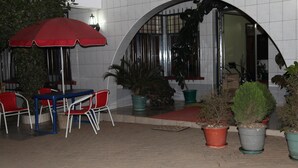Área de varanda externa com mesa, cadeiras, plantas em vasos e um guarda-sol vermelho.