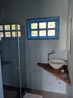 Uma casa de banho com chuveiro em cabine de vidro, uma pia branca sobre uma prateleira de madeira e uma janela com estrutura azul.