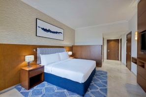 Quarto de hotel com uma cama grande, uma mesa de cabeceira com abajur, cabeceira de madeira, um quadro na parede e uma TV em uma estante de madeira.