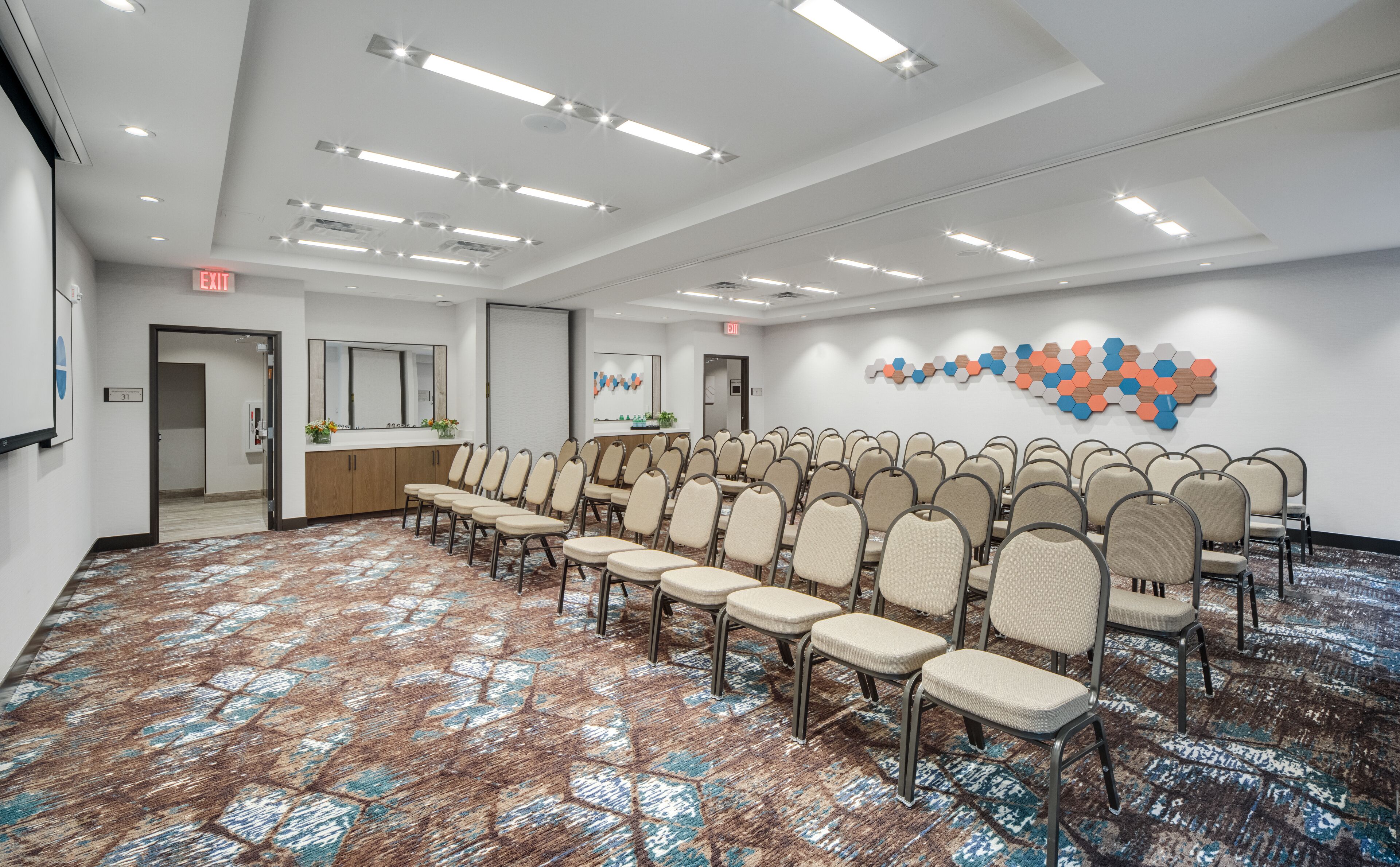 Uma sala de conferências com fileiras de cadeiras, piso carpete e um ornamento hexagonal fixado na parede, além de um sinal de saída.