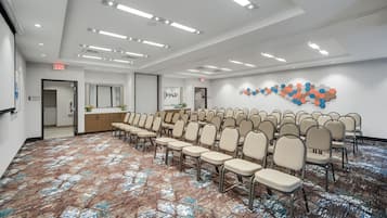 Uma sala de conferências com fileiras de cadeiras, piso carpete e um ornamento hexagonal fixado na parede, além de um sinal de saída.