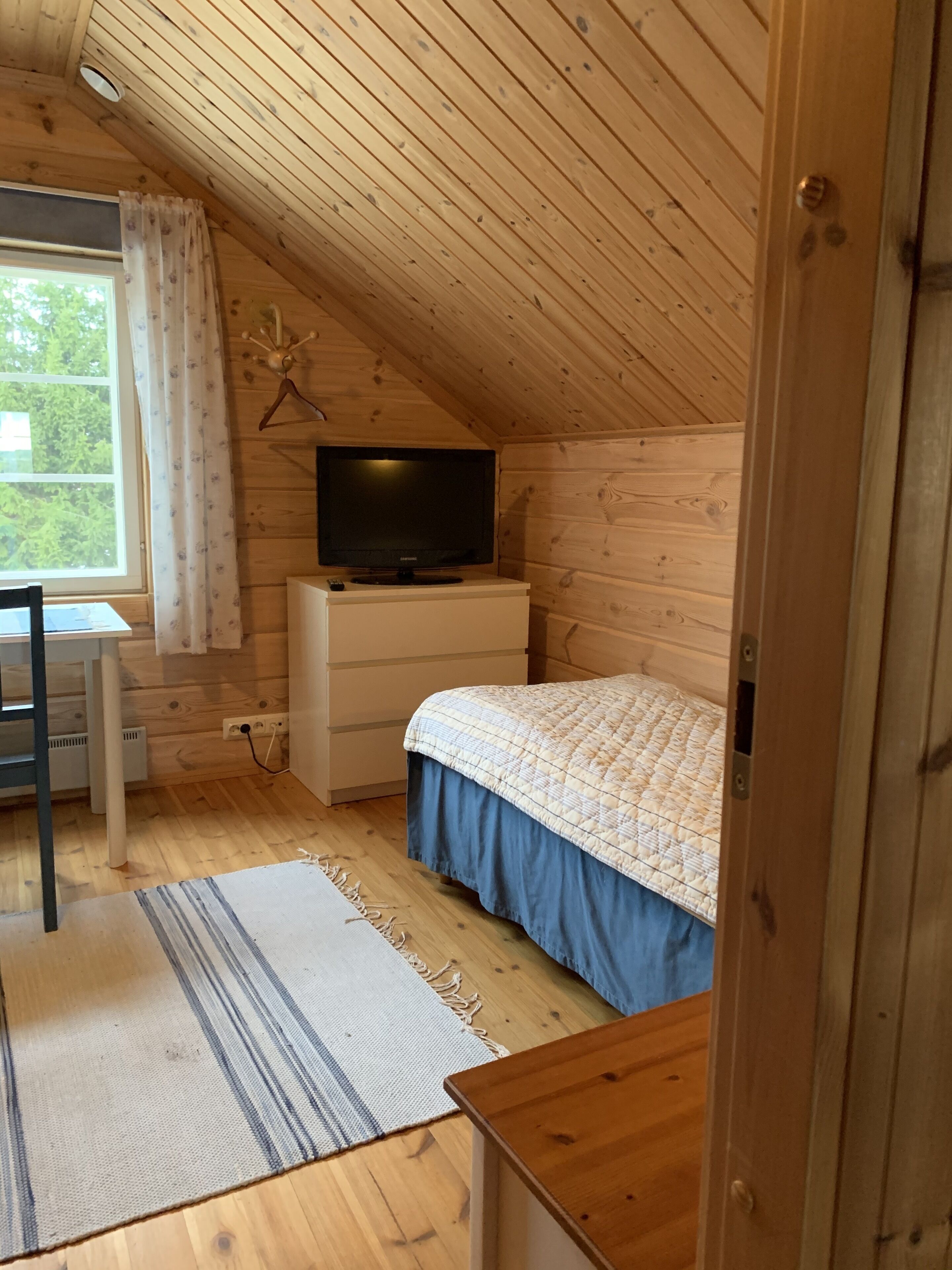 Um quarto aconchegante com teto inclinado, uma cama com colcha azul e branca, cômoda, televisão sobre suporte e janela com cortinas.