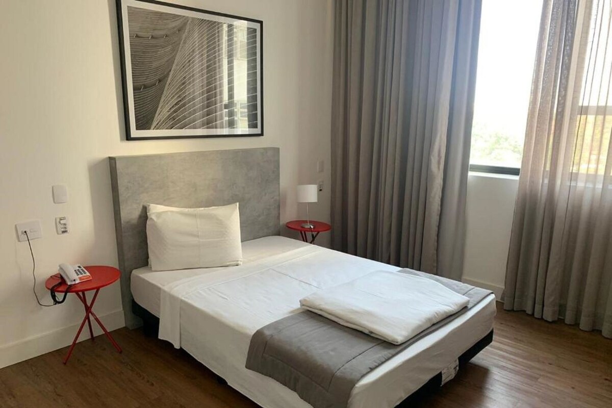 Quarto de hotel com uma cama grande, uma mesinha de cabeceira com telefone, uma mesa vermelha e uma janela com cortinas.