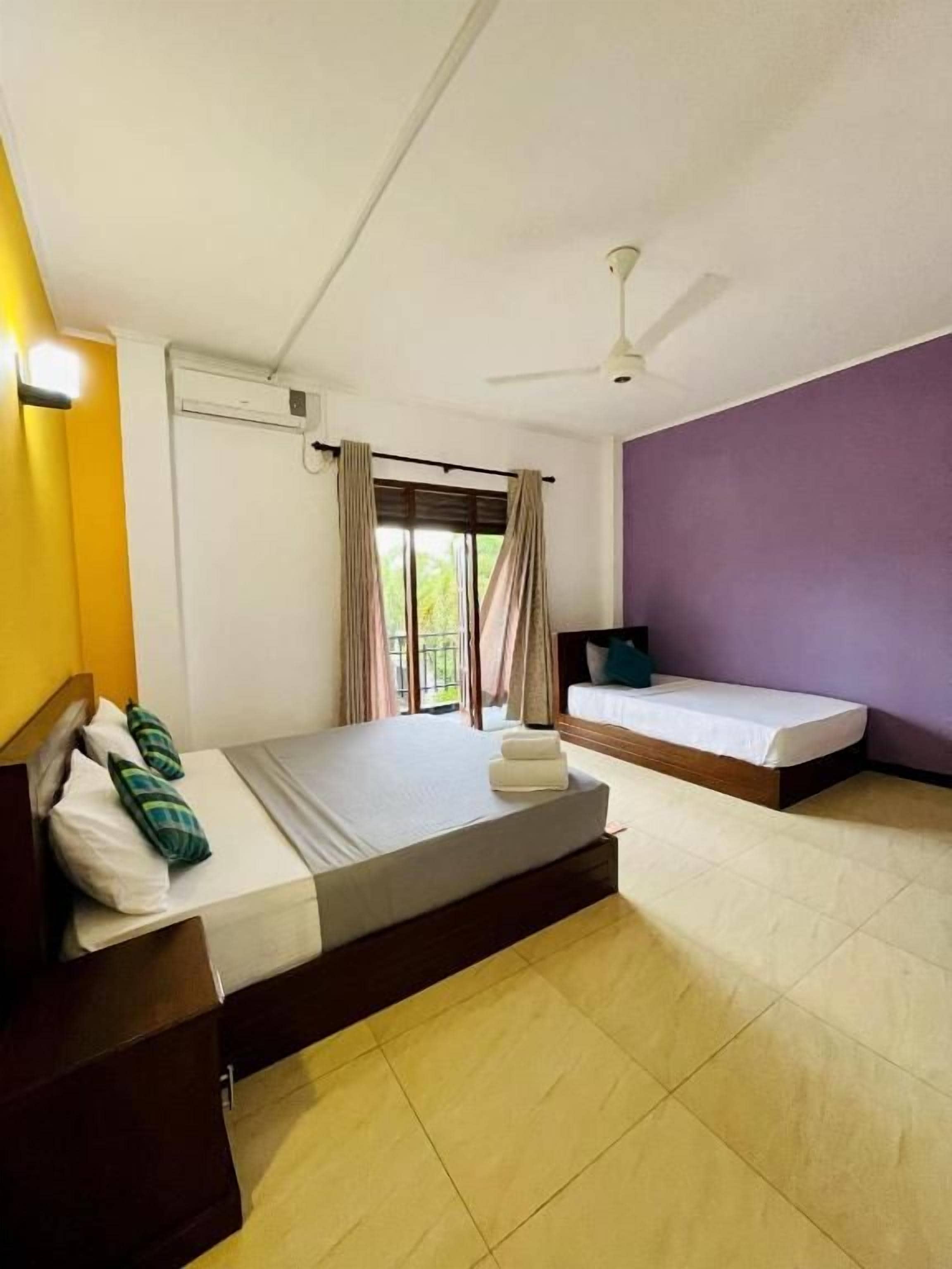 Quarto de hotel com cama, uma cama menor, ventoinha no teto e janela com cortinas.