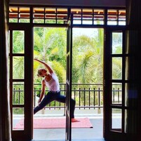 Uma pessoa fazendo uma pose de yoga em um quarto com vista para palmeiras através de uma porta.
