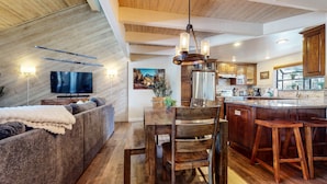 Cozinha moderna com armários de madeira, área de refeições com mesa e cadeiras de madeira, e sala de estar com sofá e televisão.