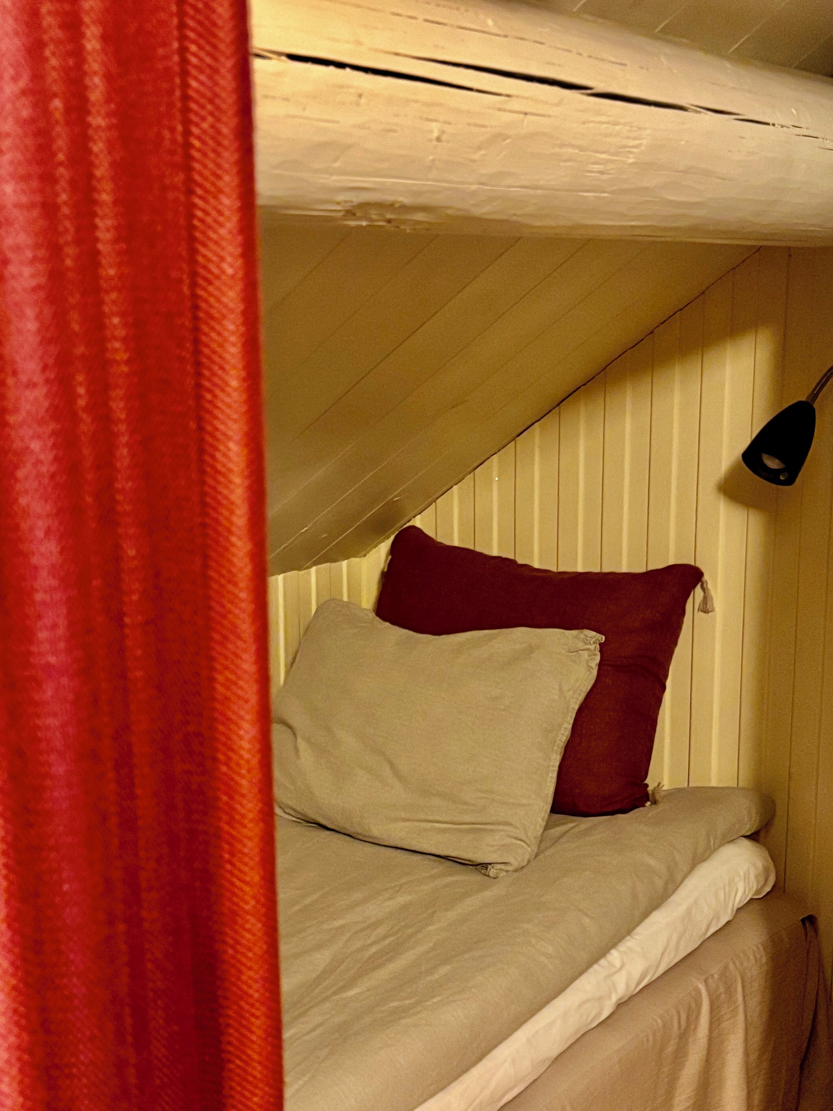 Quarto pequeno e acolhedor com uma cama de casal, cortina vermelha e teto inclinado.