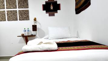 Uma cama bem arrumada com travesseiro branco e manta colorida, uma pequena mesa de cabeceira de madeira com uma garrafa de água e uma vela, e uma parede decorada com arte em padrão.