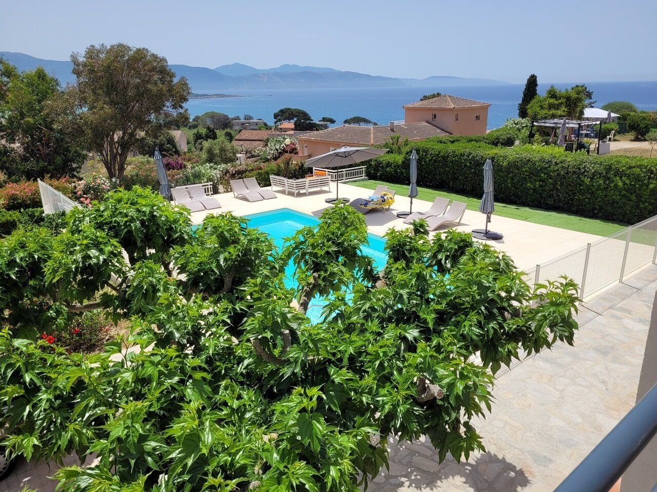 Área de piscina com espreguiçadeiras e guarda-sóis, cercada por vegetação exuberante e vista para o mar e montanhas.